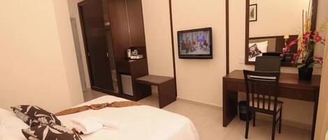 Quarto de hotel com cama, escrivaninha, cadeira, guarda-roupa e televisão.