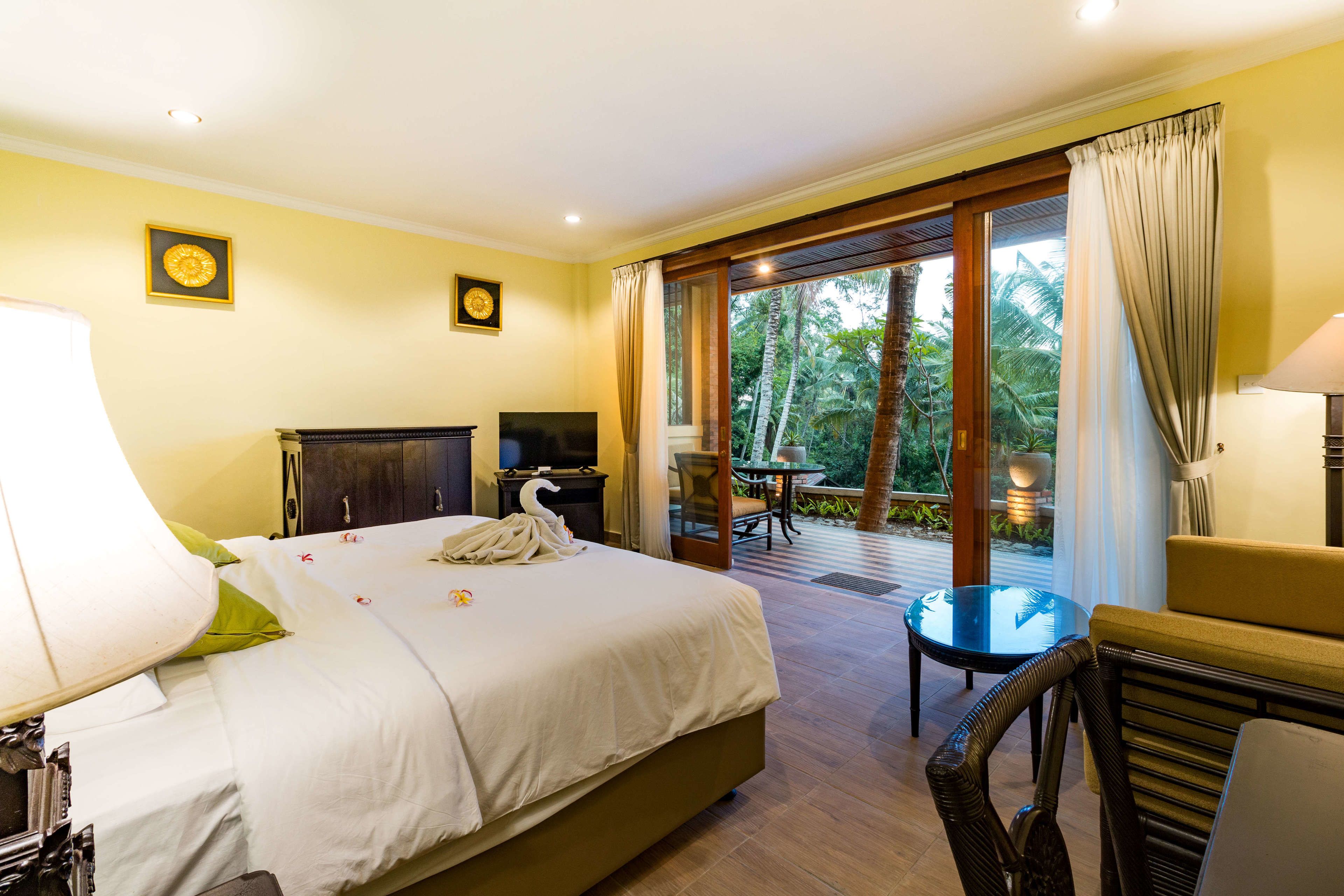 Quarto de hotel com cama, televisão, lareira, varanda com mesa e cadeiras, e vista para palmeiras.