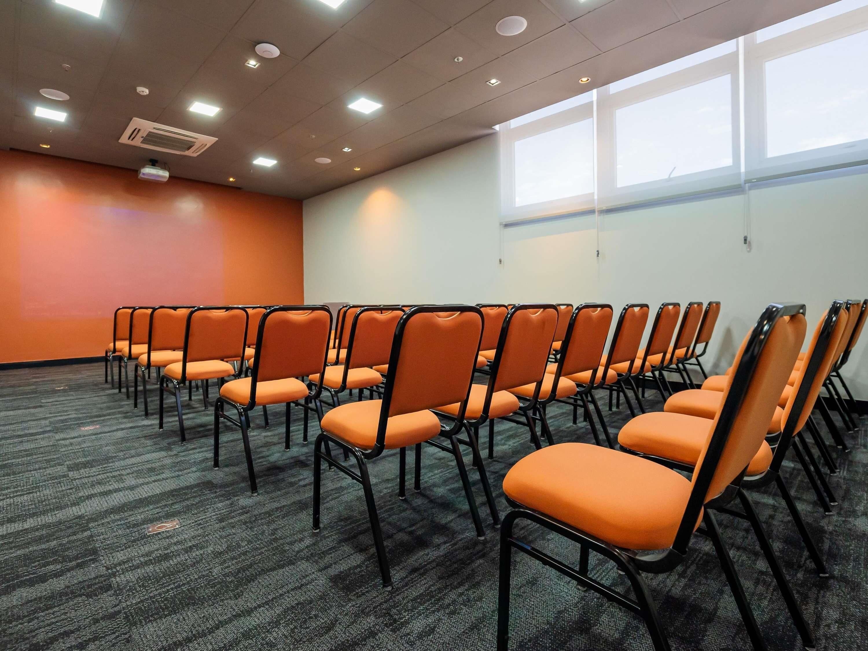 Uma sala de conferências com fileiras de cadeiras laranja, piso coberto de carpete, luminárias no teto e janelas.