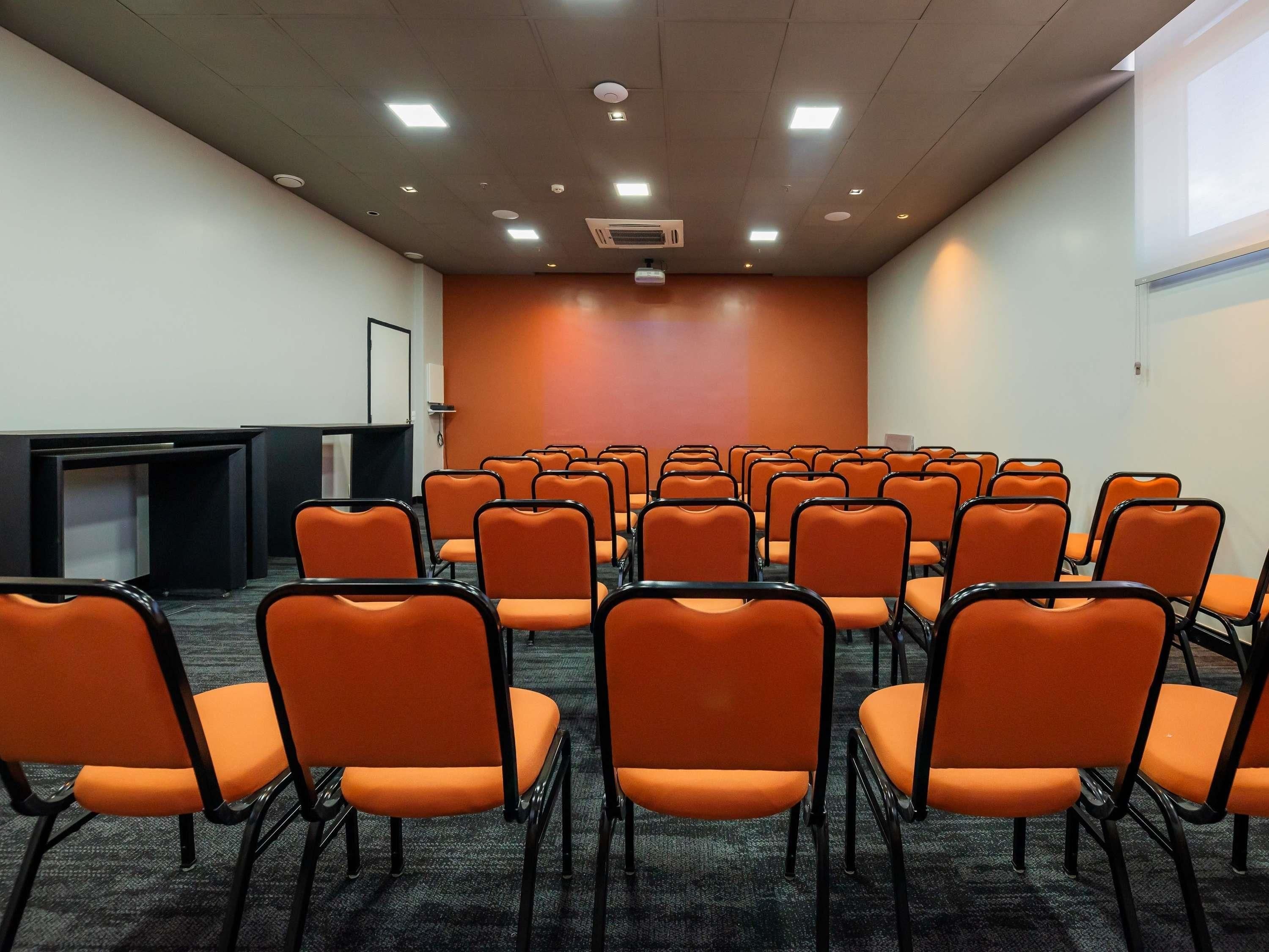 Uma sala de conferências com fileiras de cadeiras laranjas, uma tela de projetor e um ecrã de apresentação.