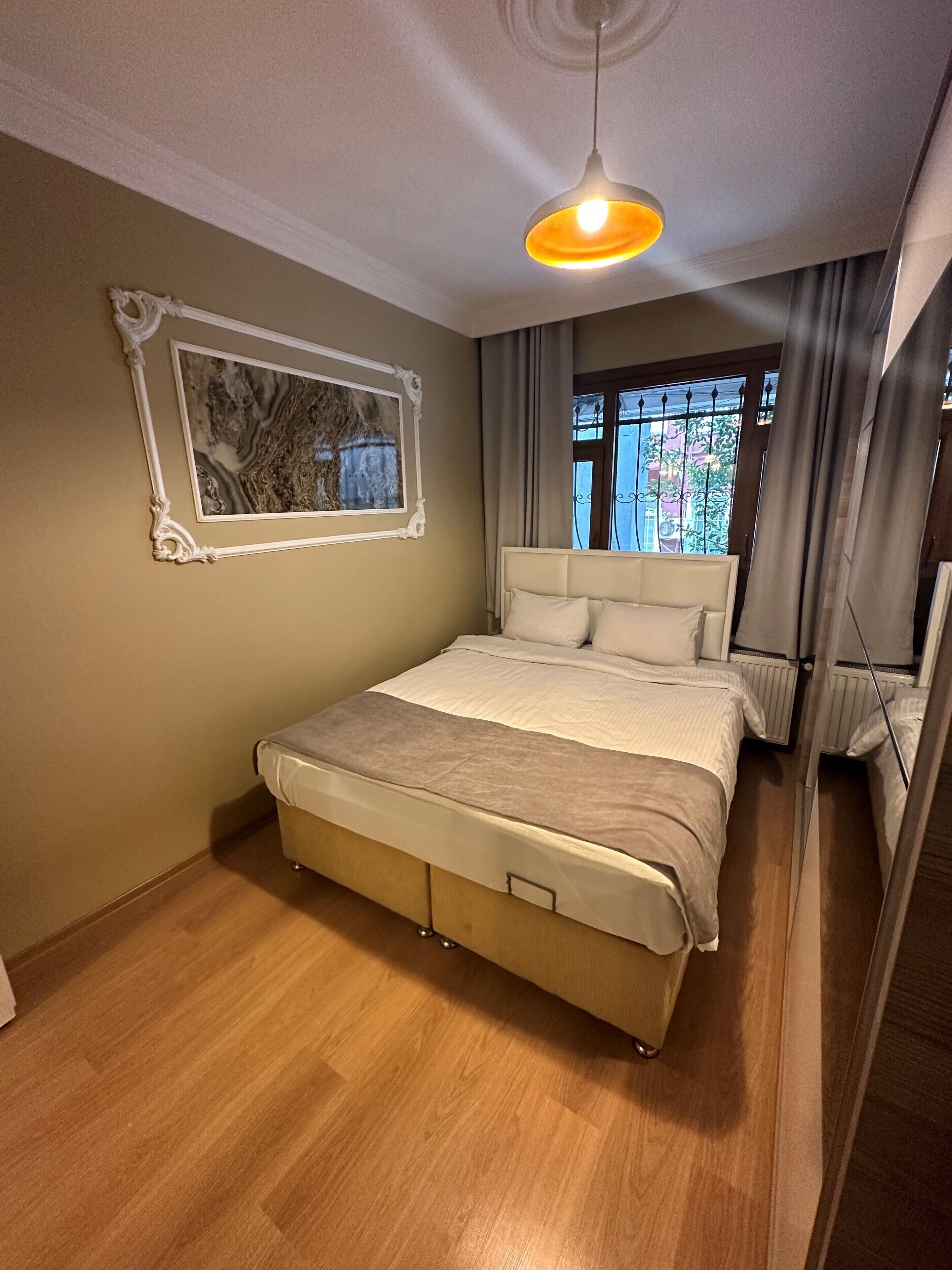 Um quarto com uma cama grande, uma luminária no teto, uma obra de arte emoldurada e uma janela com cortinas.