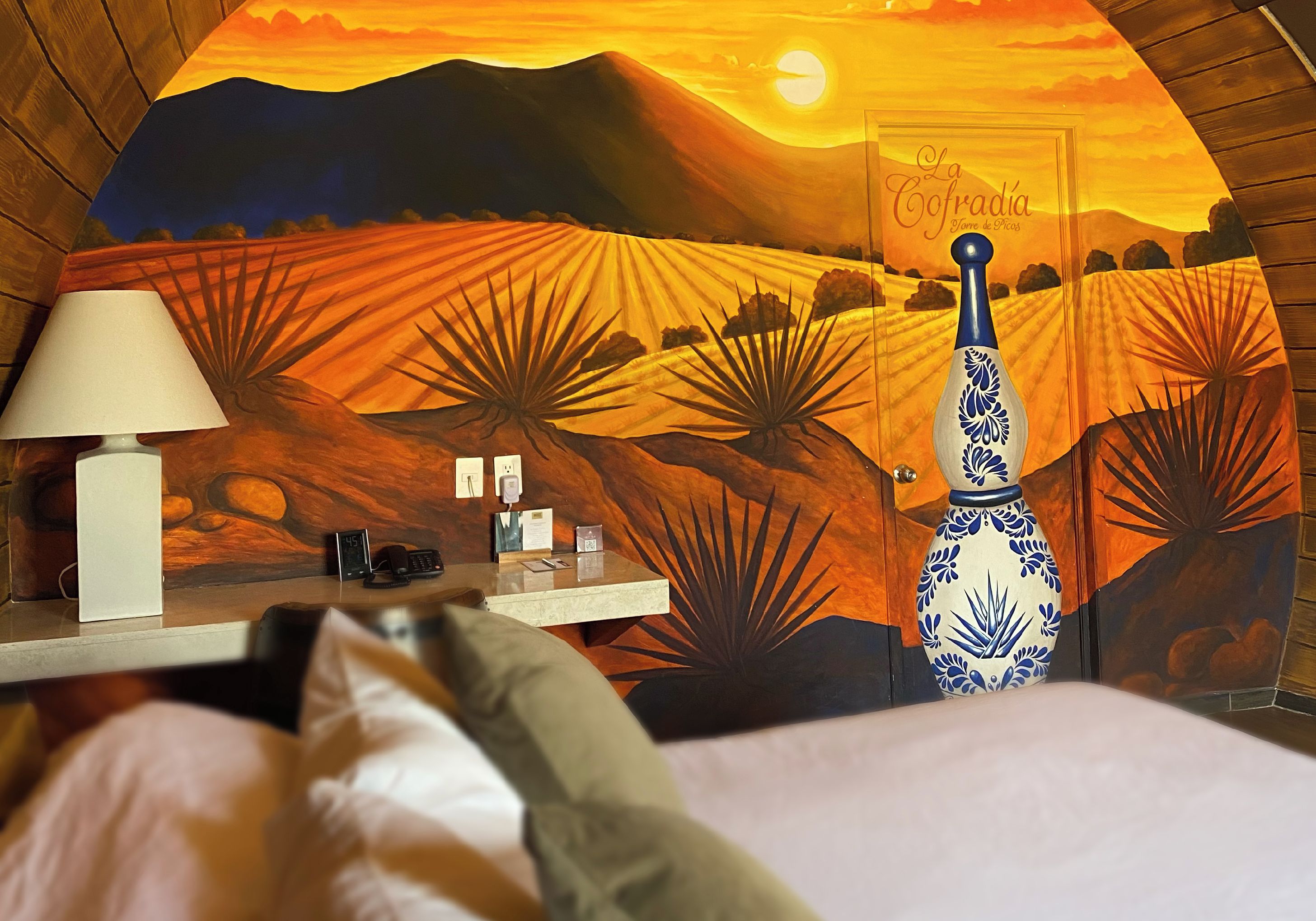 Quarto de hotel com um mural de paisagem ao pôr do sol, uma mesa de cabeceira com abajur, um vaso e um relógio.