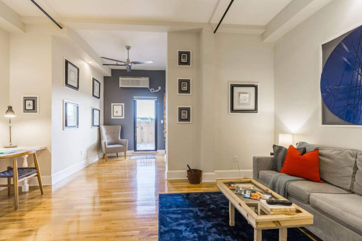 Uma sala de estar com piso de madeira, um sofá cinza com almofadas laranja, uma mesa de centro e quadros nas paredes.