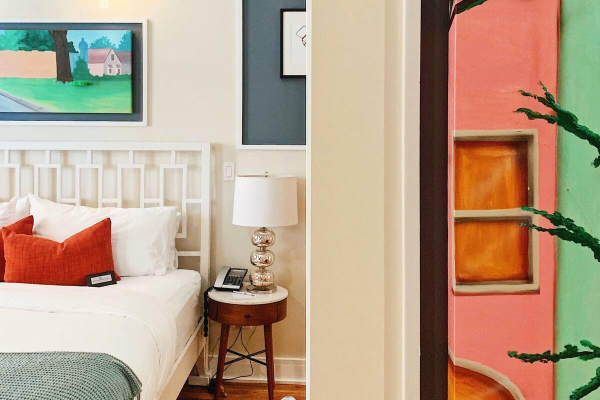 Quarto de hotel com cama, mesa de cabeceira com abajur, quadro na parede e janela com cortinas.