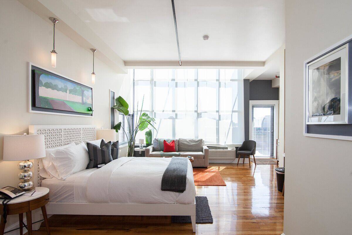 Quarto moderno com uma cama grande, um sofá e uma escrivaninha. Há janelas amplas e vista para o exterior.