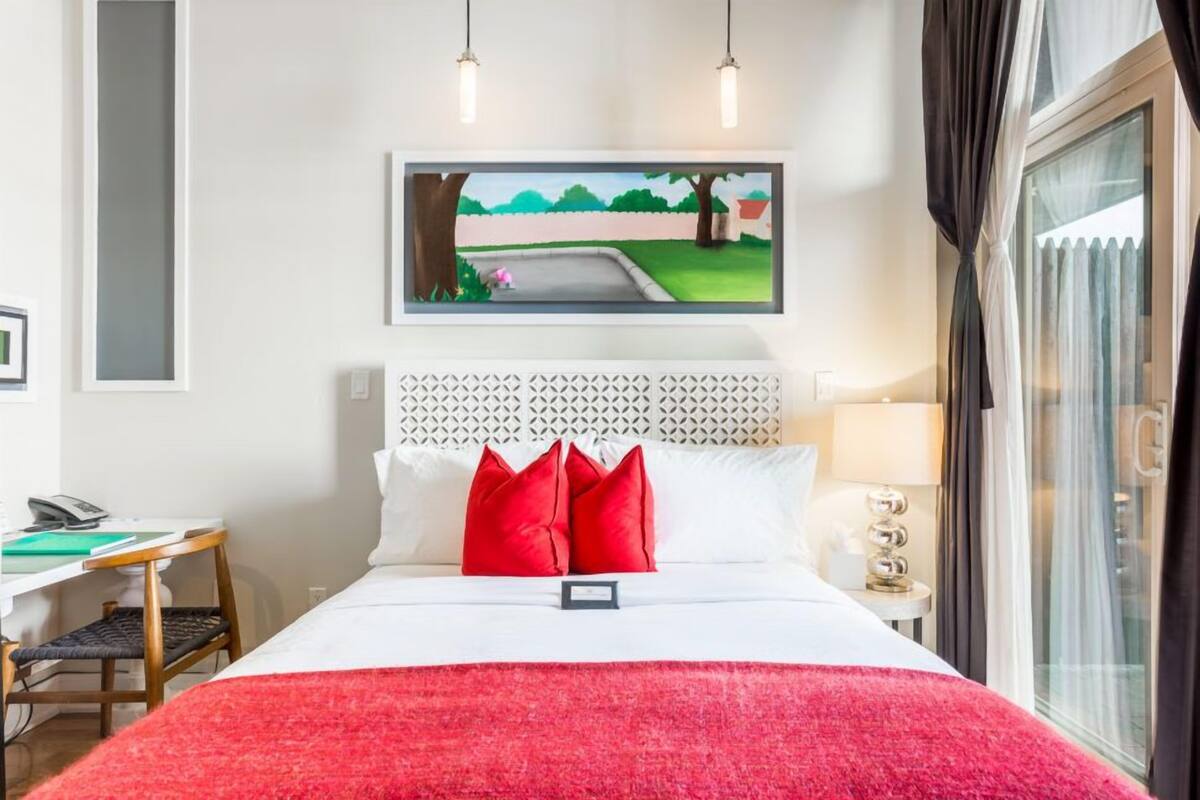 Quarto de hotel com cama, escrivaninha, cadeira, um quadro na parede e janela com cortinas.