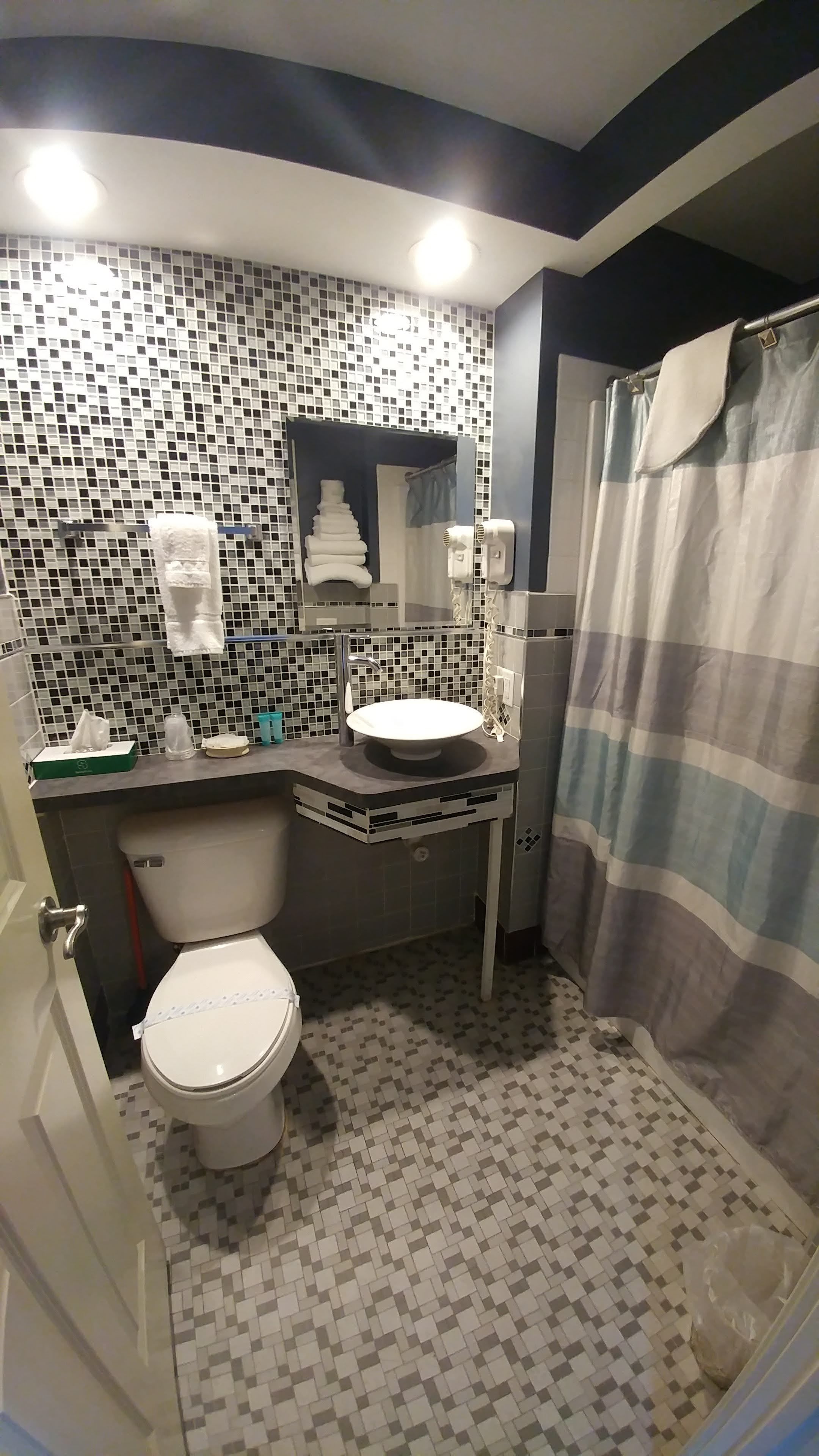 Um banheiro com piso ladrilhado, vaso sanitário branco, pia com espelho acima e cortina de chuveiro.