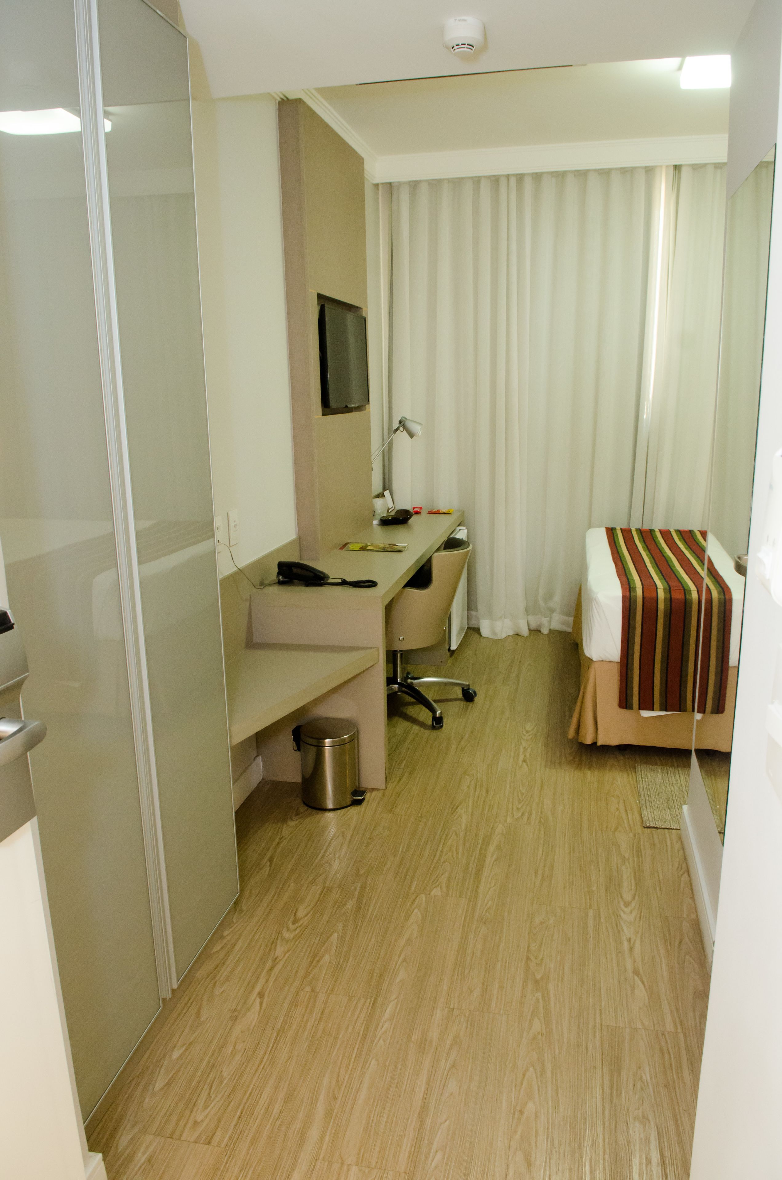 Quarto de hotel pequeno com escrivaninha, cadeira e cama.