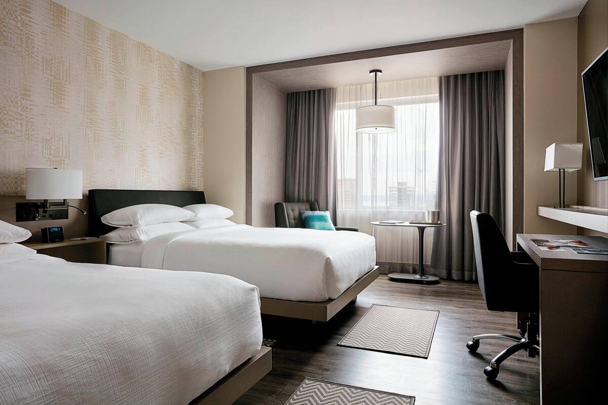 Quarto de hotel com duas camas, uma escrivaninha, uma cadeira, um abajur e uma televisão.
