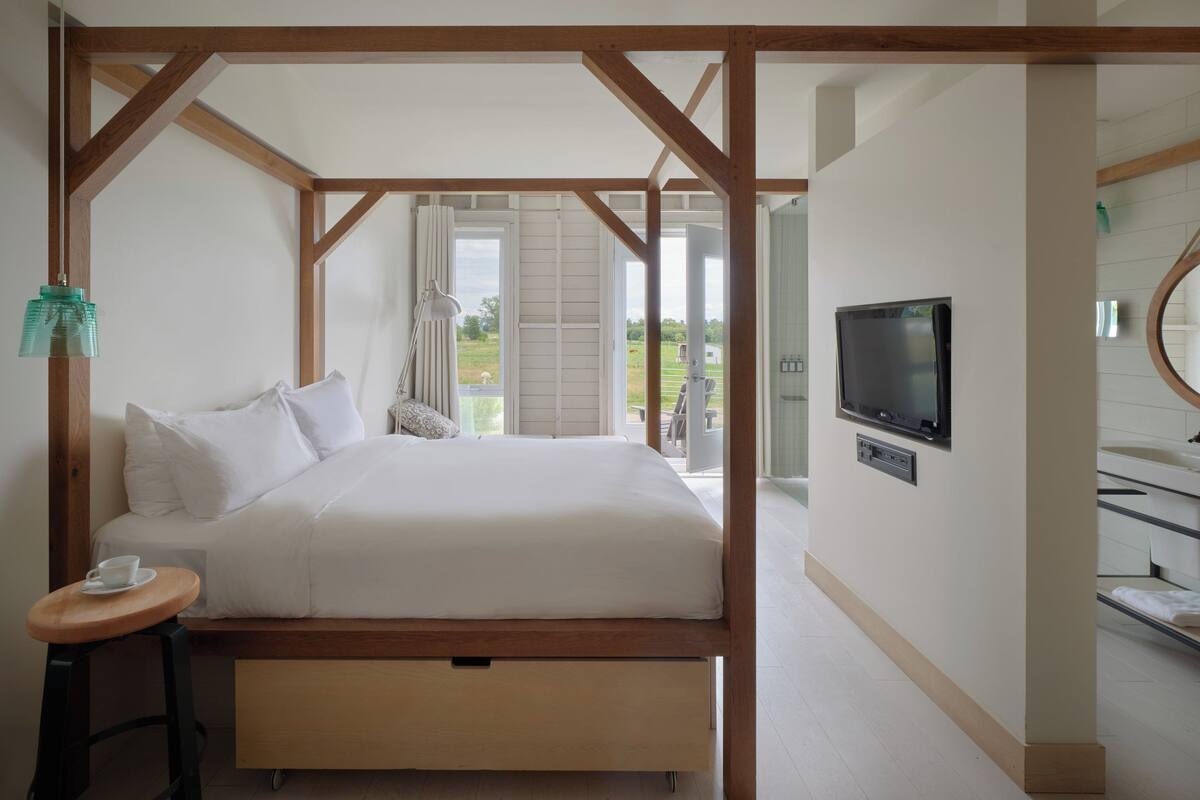 Quarto com cama, televisão, secretária com cadeira e janela com cortinas.