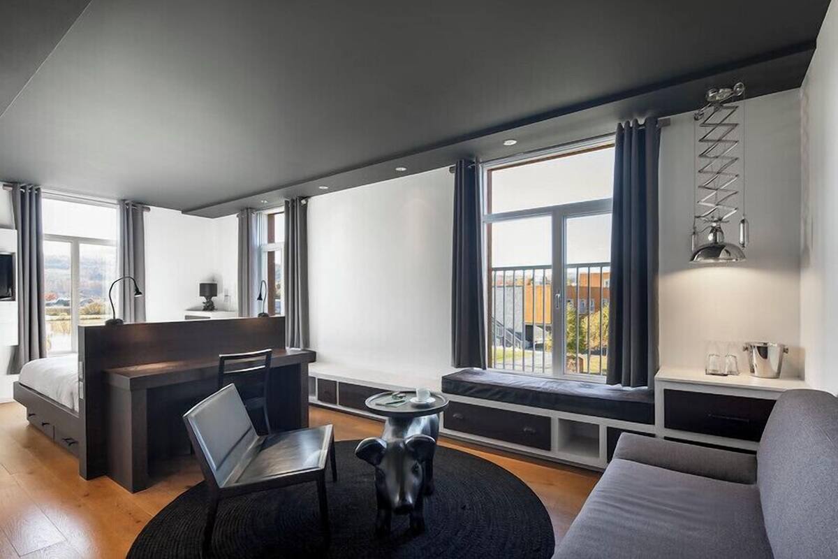 Quarto moderno com cama, escrivaninha, poltrona e sofá, janelas amplas e vista para a cidade.