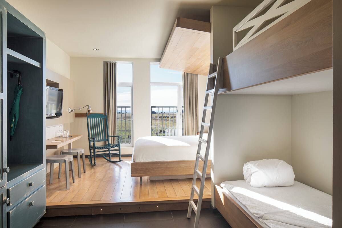 Quarto de hotel compacto com beliche, escrivaninha com cadeira, televisão e vista para o exterior.