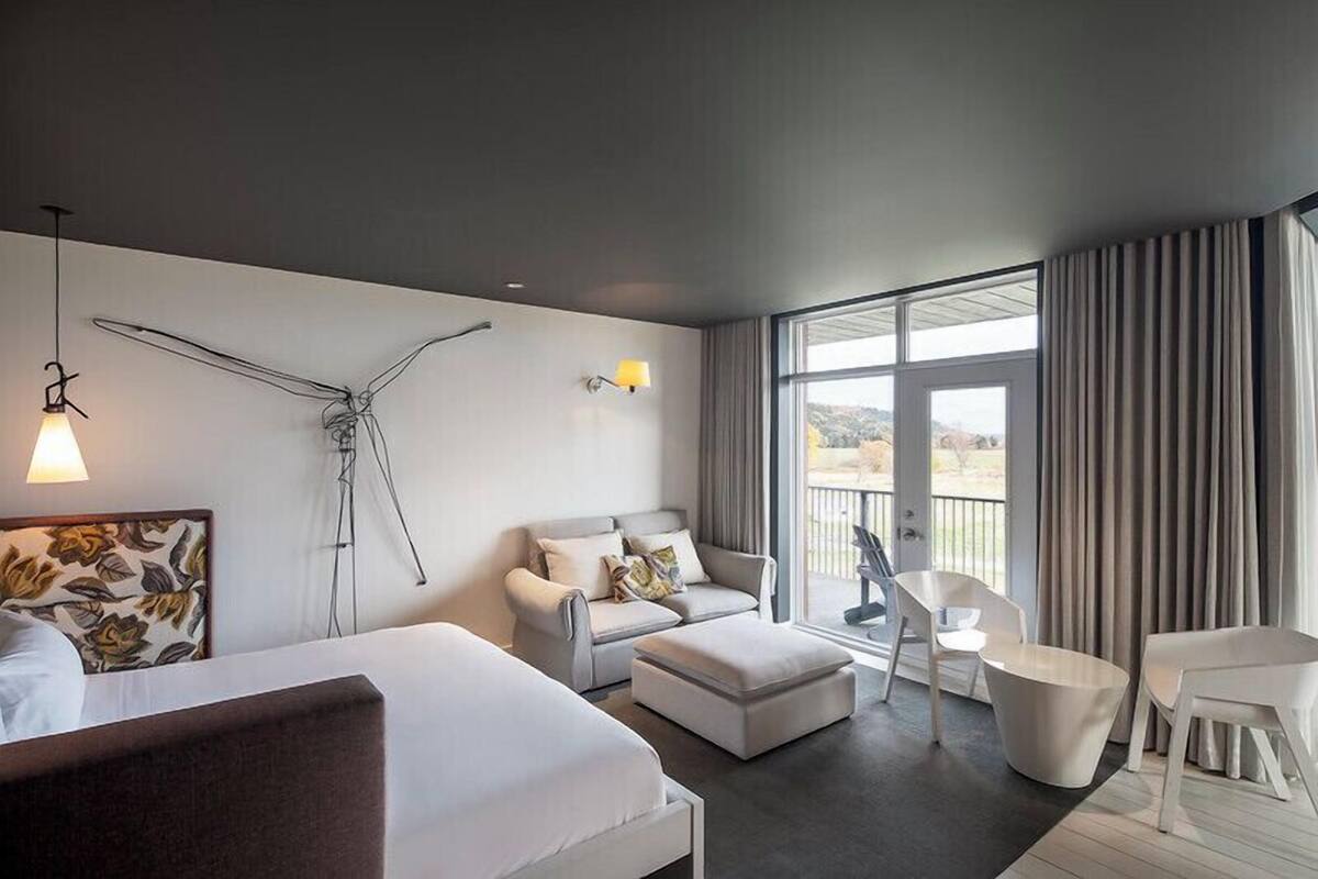 Quarto de hotel moderno com uma cama grande, um sofá, uma mesinha e uma varanda com cortinas.