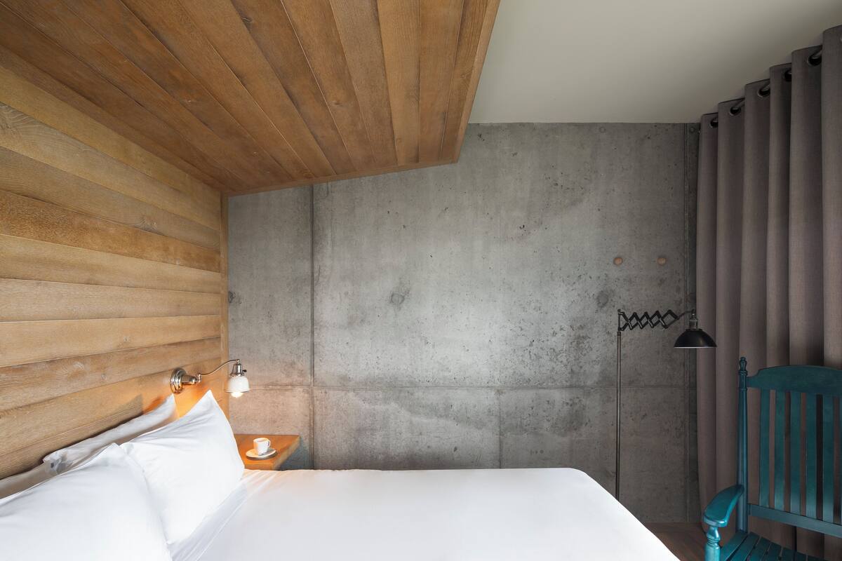 Quarto com cabeceira de madeira, cama com roupa de cama branca, mesa de cabeceira com luminária e uma cadeira azul.