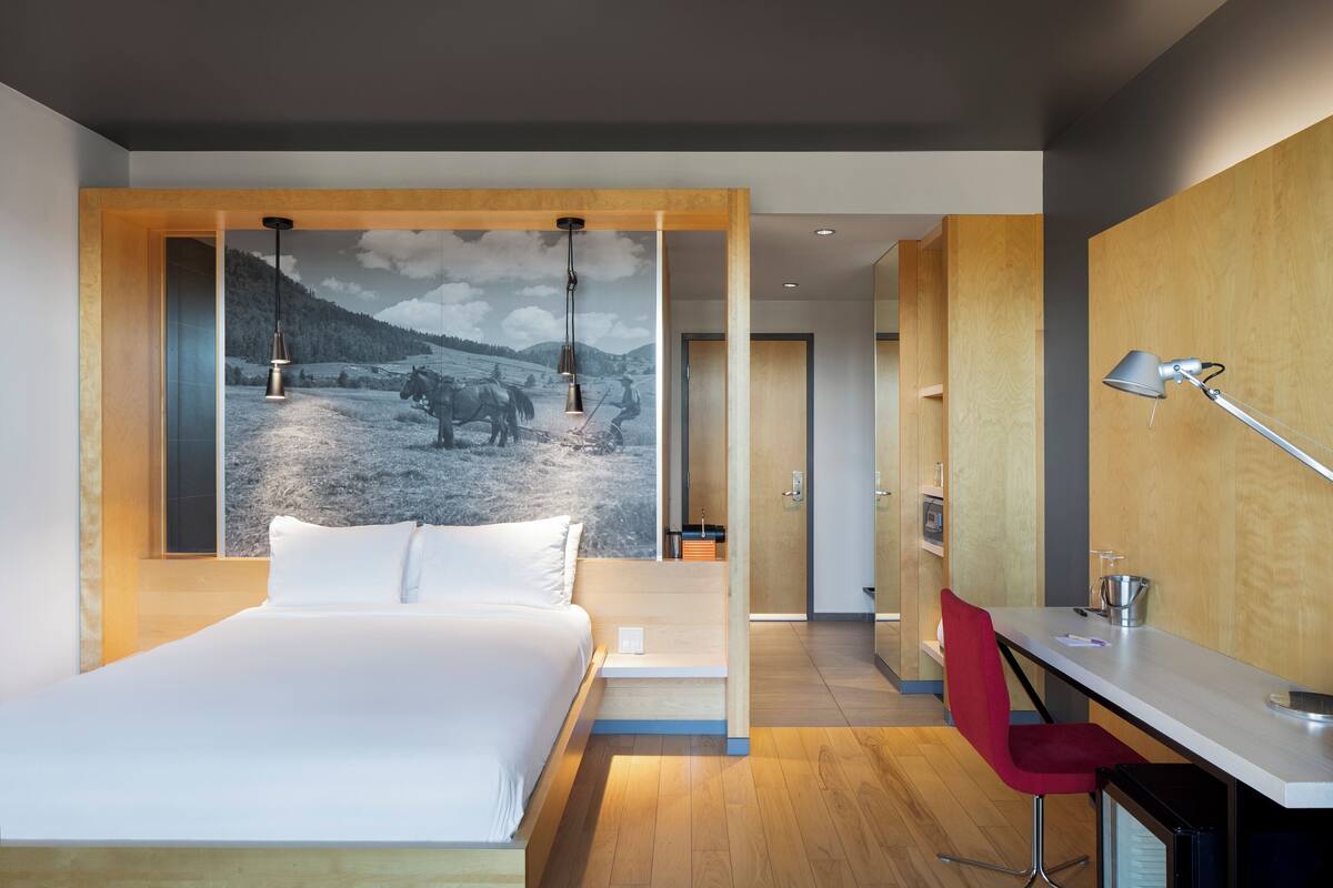 Quarto de hotel moderno com uma cama grande, uma escrivaninha e uma cadeira vermelha. Há um painel de madeira com um mural de uma paisagem montanhosa.
