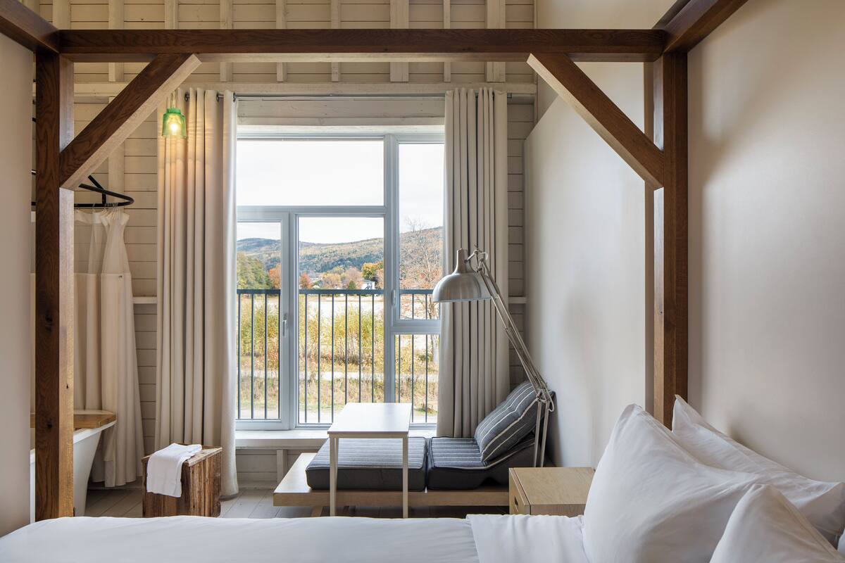 Quarto com cama, um banco com almofadas, mesa, luminária e janela com cortinas.