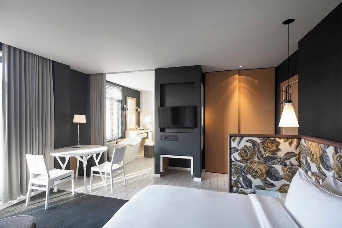 Quarto de hotel com cama, escrivaninha, cadeira, televisão e janela com cortinas.