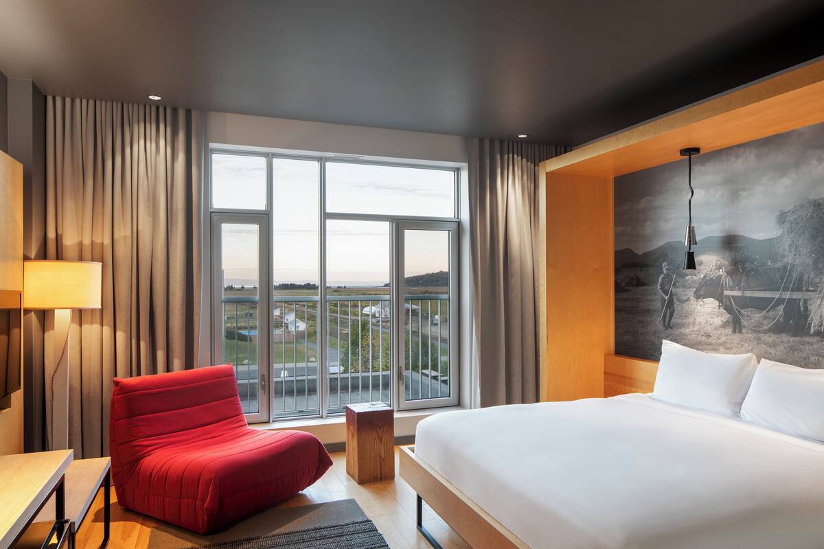 Quarto de hotel moderno com uma cama grande, um sofá vermelho, uma escrivaninha e vista para o campo através de amplas janelas.