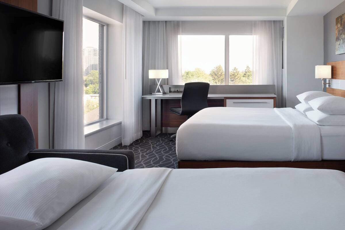 Quarto de hotel com duas camas, uma escrivaninha, uma cadeira, televisão e janela com vista para edifícios e árvores.