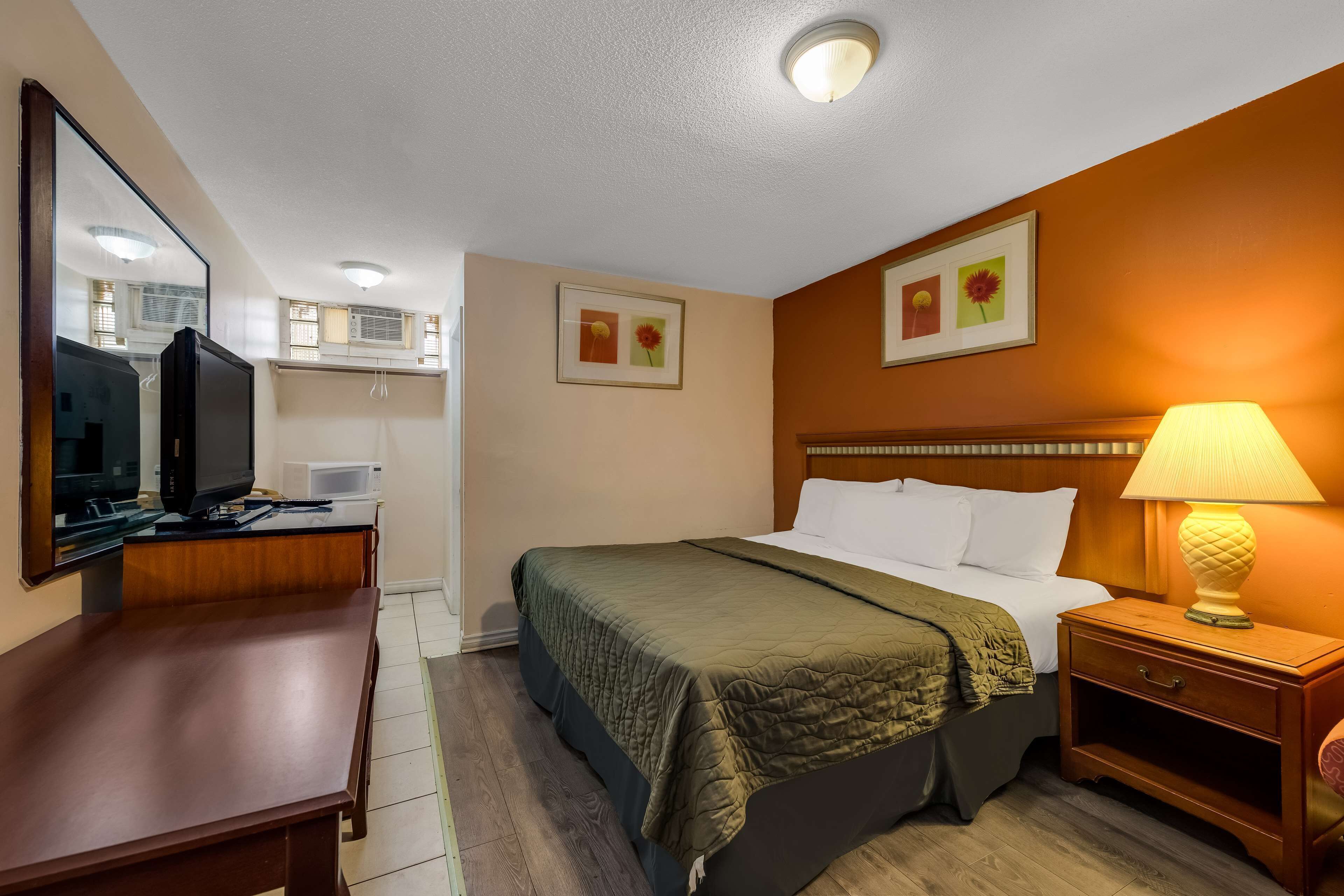 Quarto de hotel com cama, mesa de cabeceira, luminária, televisão e escrivaninha com cadeira.