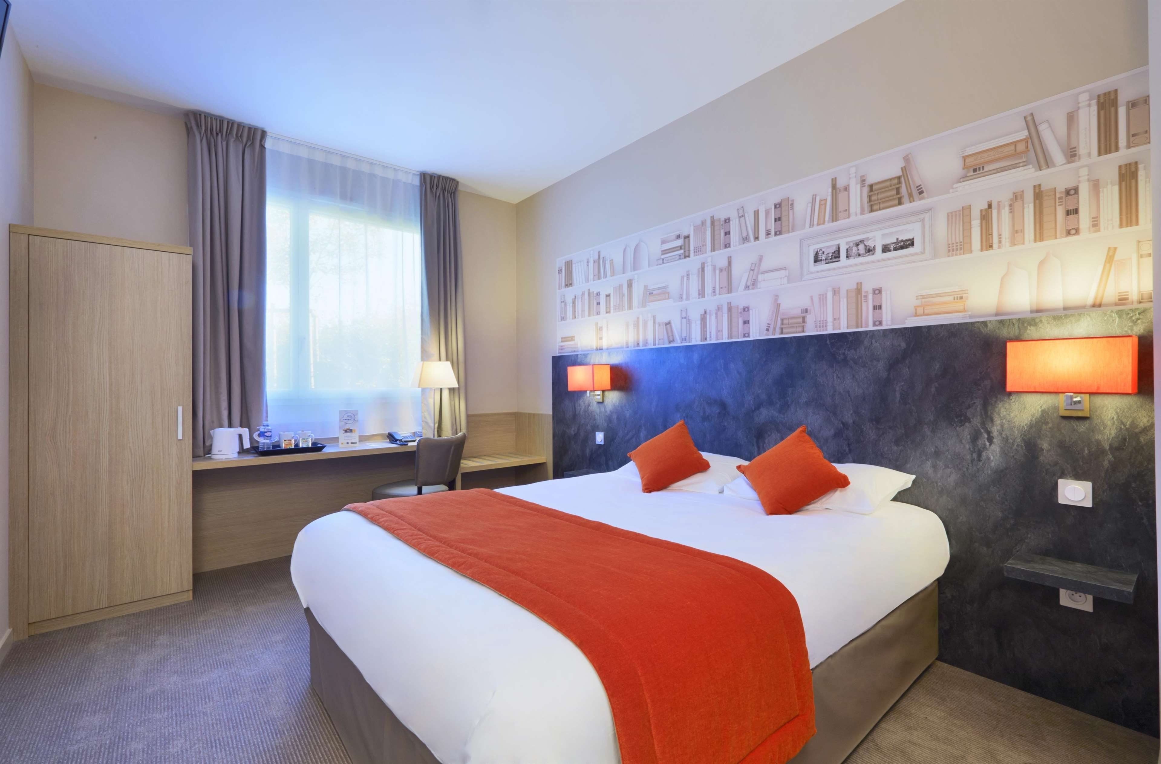 Quarto de hotel com uma cama grande, uma escrivaninha com cadeira, um guarda-roupa e uma parede com quadros.