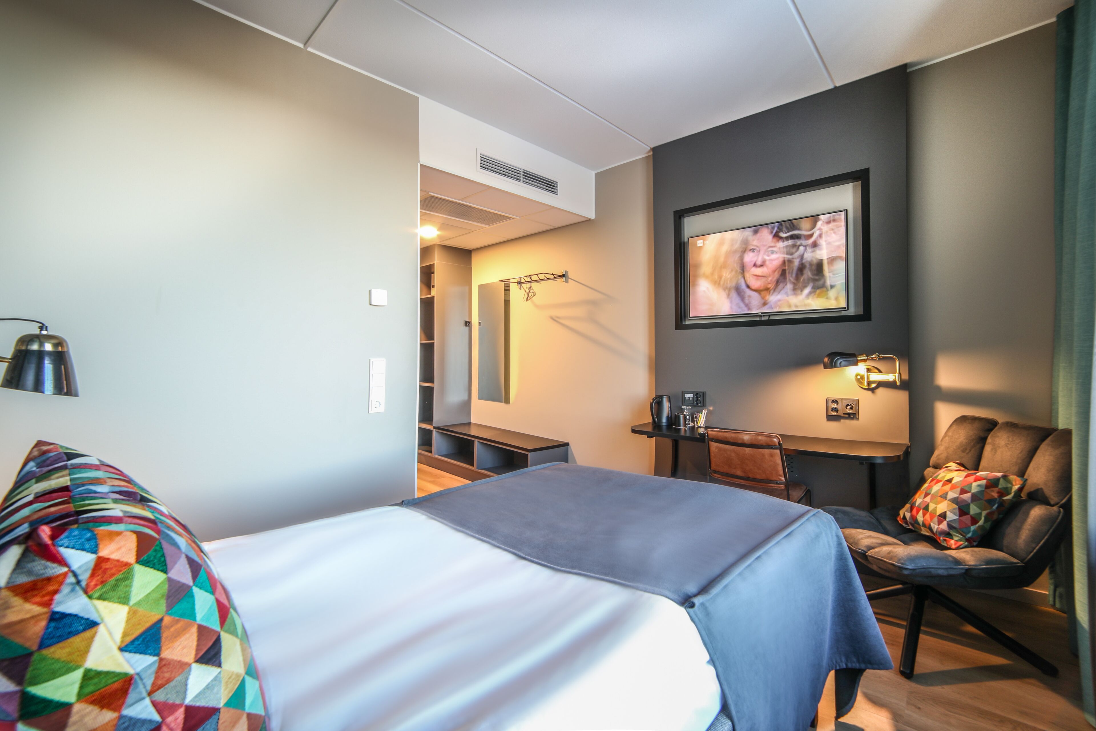 Quarto de hotel com cama, escrivaninha, cadeira e televisão.