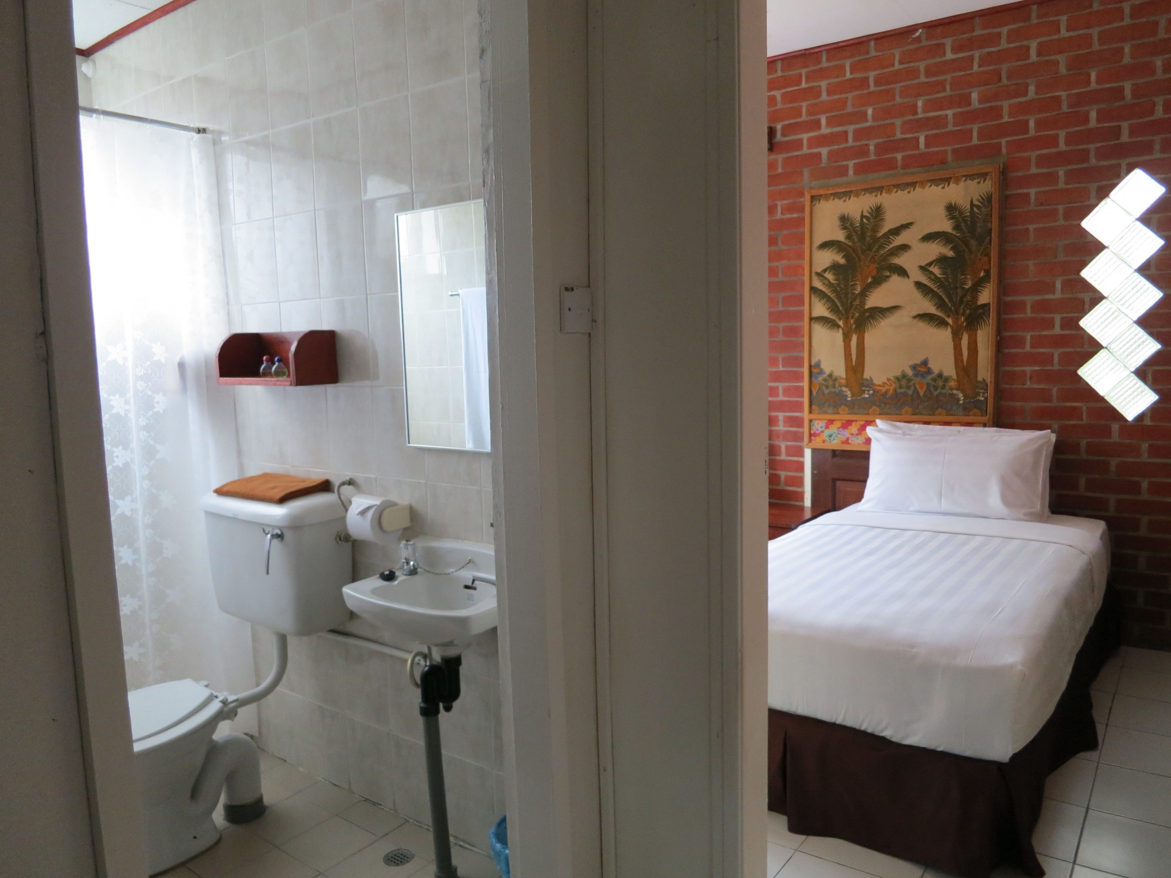 Casa de banho com sanita, lavatório e chuveiro. Um quarto adjacente apresenta uma cama e um quadro na parede.