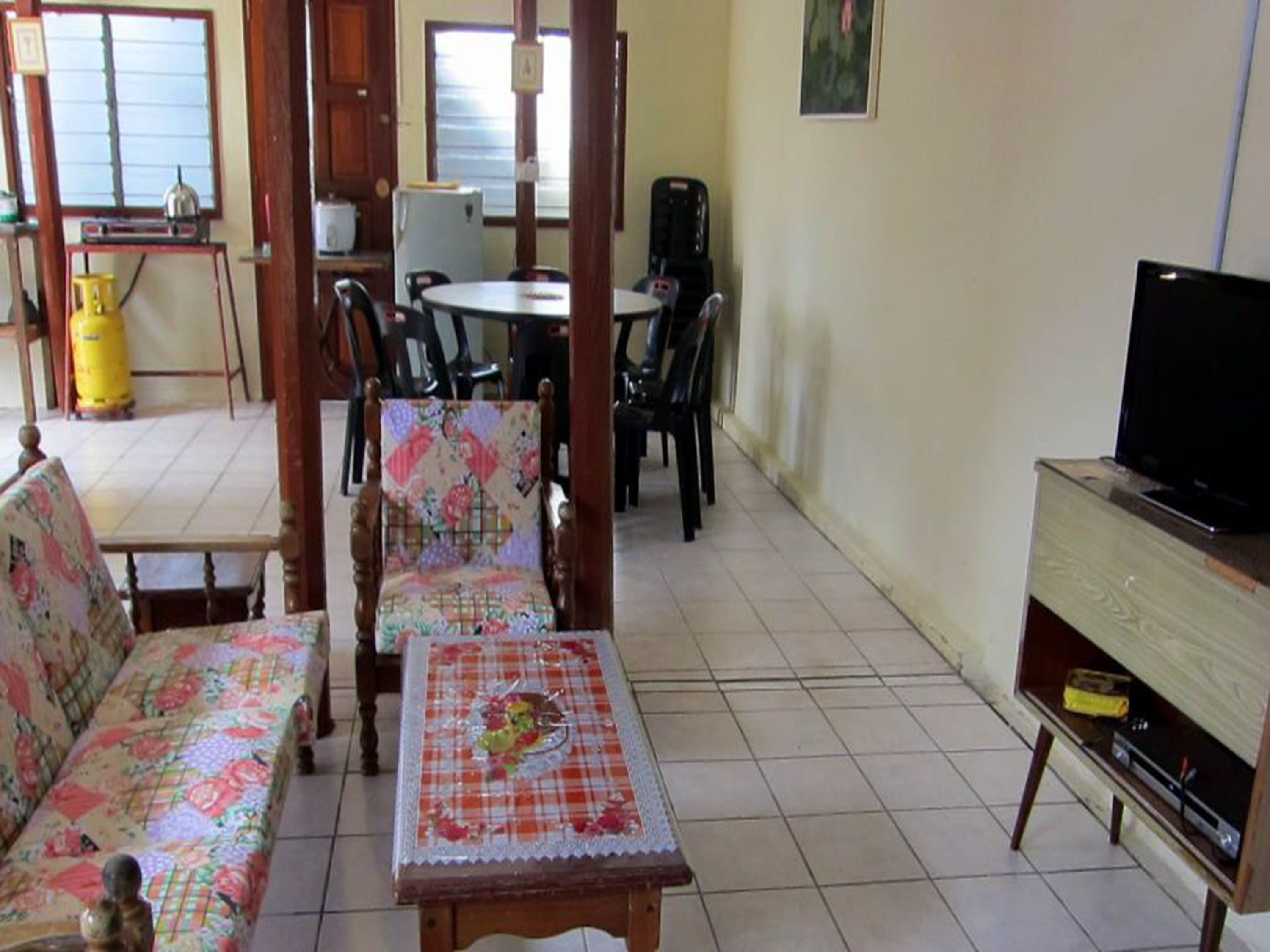 Uma sala de estar com sofá, uma mesa pequena com toalha de mesa, televisão sobre um suporte de madeira, e um espaço de refeições com cadeiras e mesa.