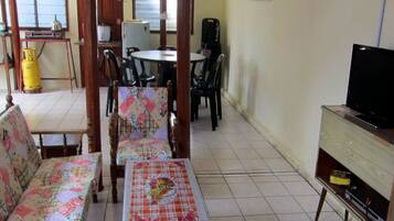 Uma sala de estar com sofá, uma mesa pequena com toalha de mesa, televisão sobre um suporte de madeira, e um espaço de refeições com cadeiras e mesa.