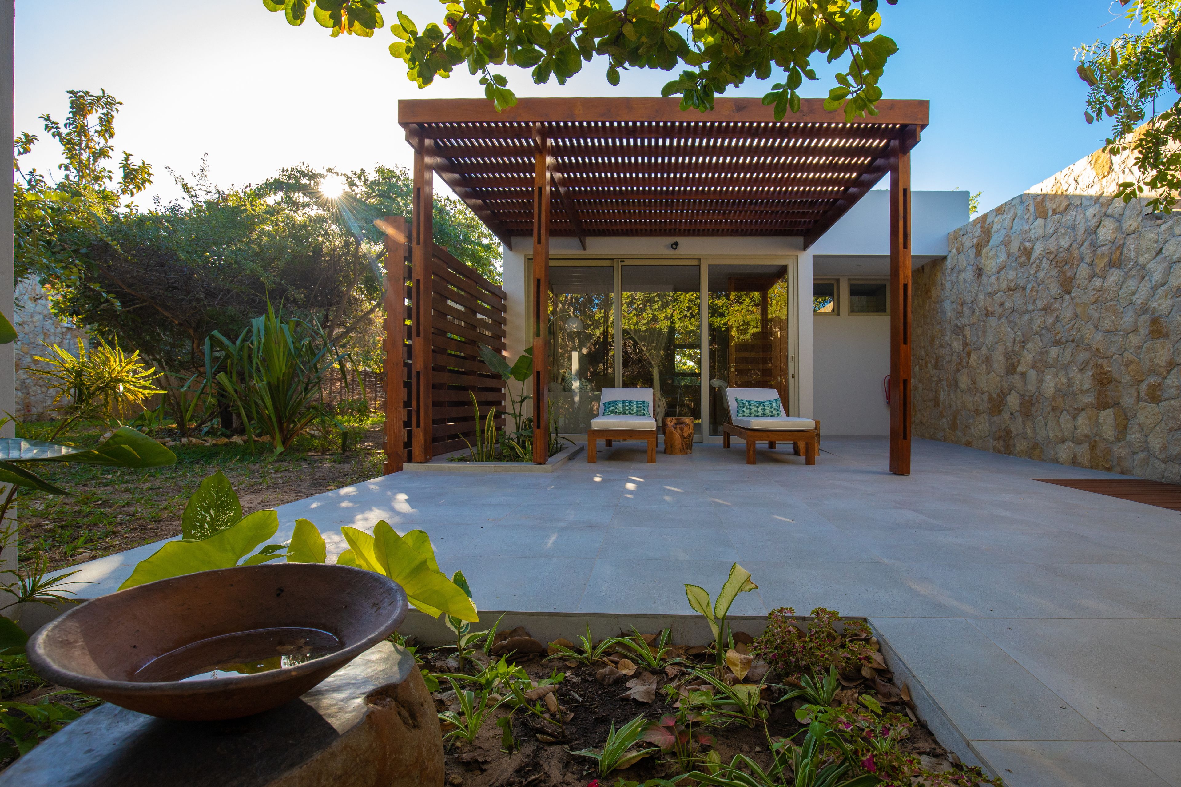 Uma área de estar coberta ao ar livre, com duas cadeiras e uma mesa, cercada por plantas e um muro de pedra.