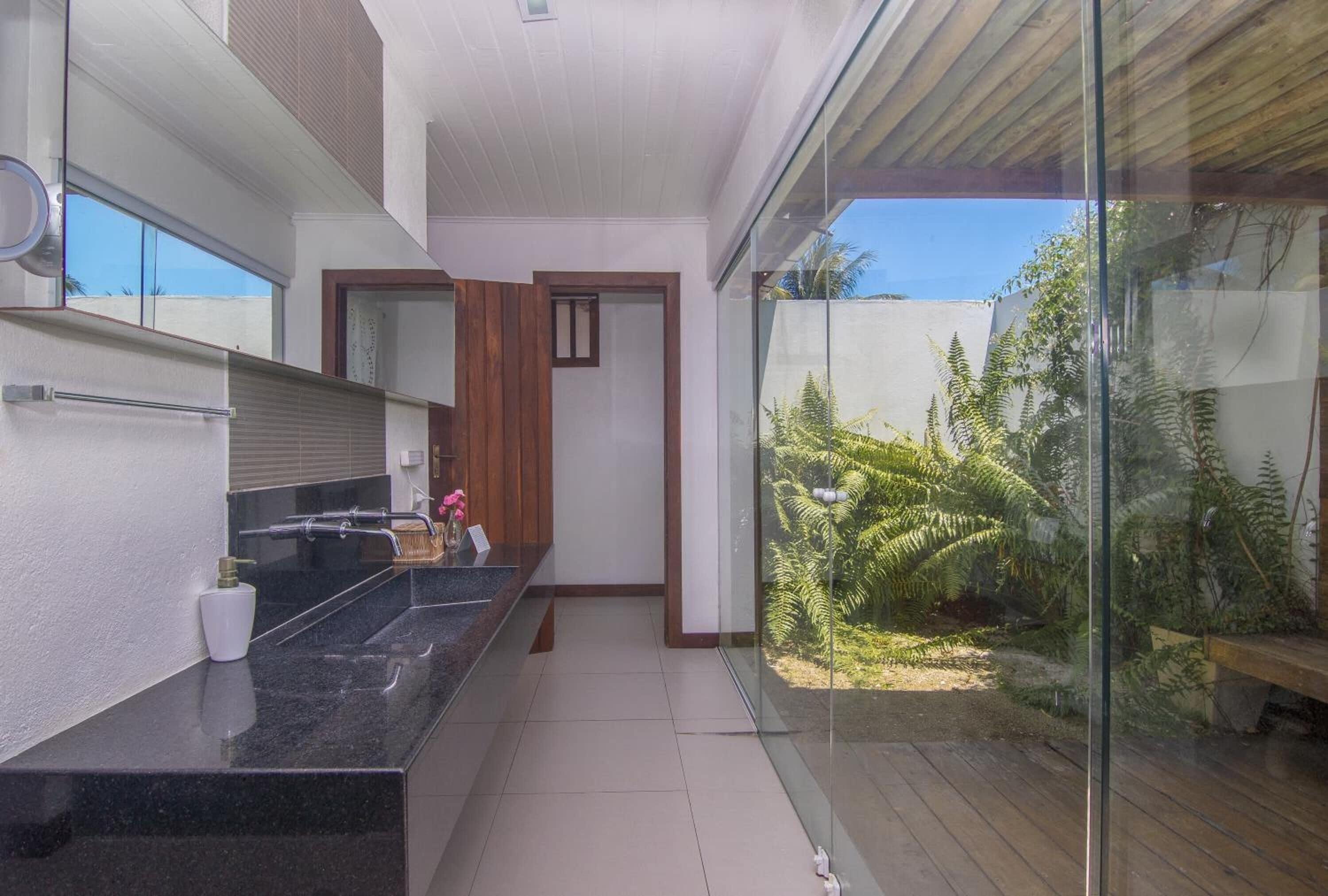 Casa de banho moderna com bancada preta, lavatório branco e cabine de vidro que dá para um jardim com samambaias.
