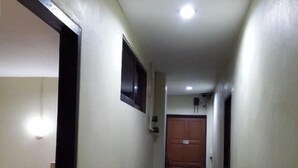 Um corredor estreito com uma cama à esquerda, uma porta no final e um aparelho de luz na parede.