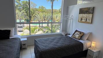 Quarto com uma janela grande, cama com colcha estampada, mesa de cabeceira com abajur e uma obra de arte na parede.