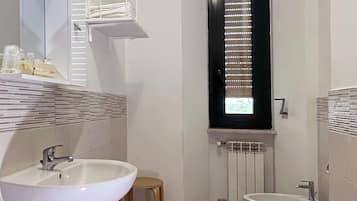 Um banheiro com pia branca, dois vasos sanitários, uma janela com persianas e um radiador.