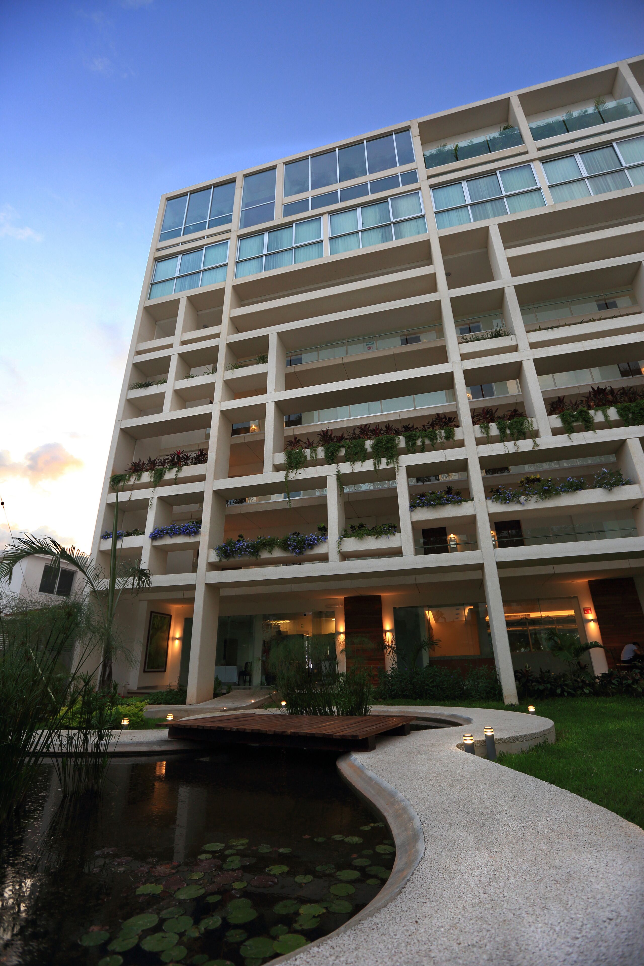 Um hotel com vários andares, um pátio central, um pequeno espelho d'água e varandas com plantas.