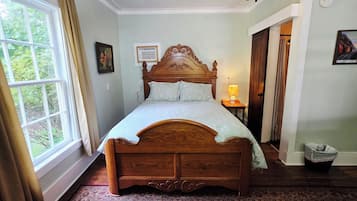 Quarto com cama grande, janela com cortinas, cabeceira de madeira, mesa de cabeceira com abajur e porta para outro cômodo.