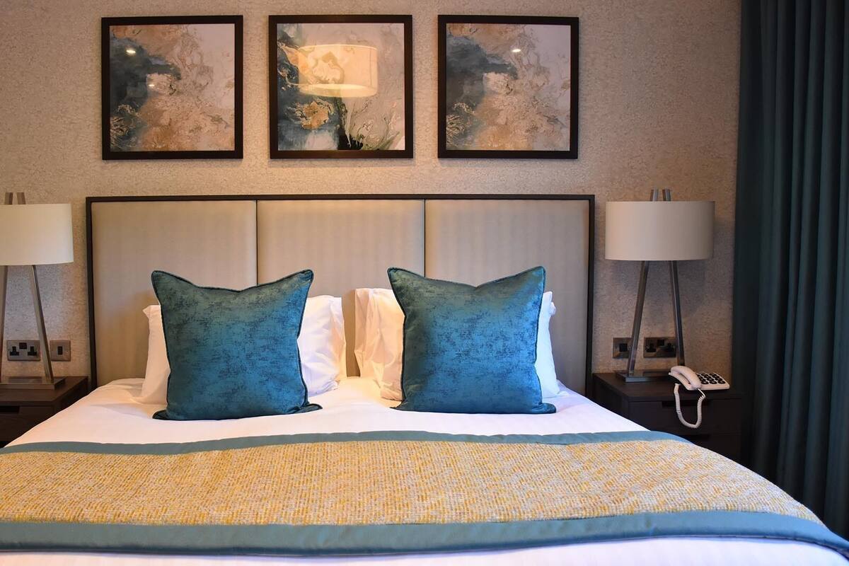 Quarto de hotel com uma cama grande, duas almofadas, mesa de cabeceira com abajur, telefone e três quadros na parede.