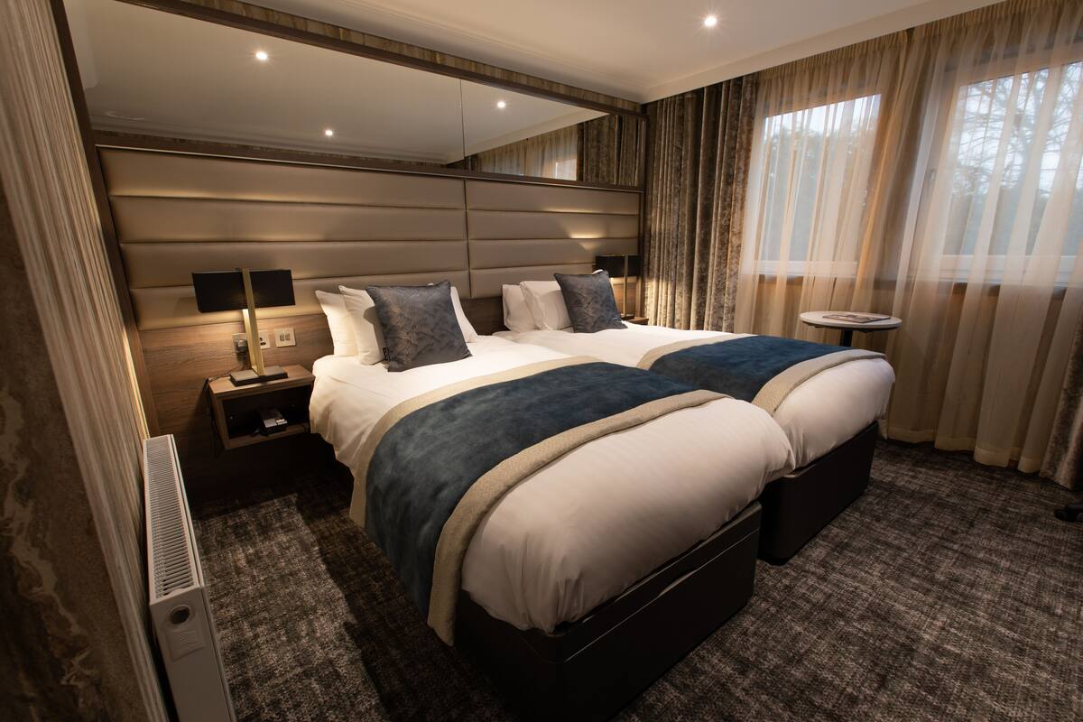 Quarto de hotel com duas camas, uma mesinha de cabeceira com abajur, uma mesa redonda e uma janela grande com cortinas.