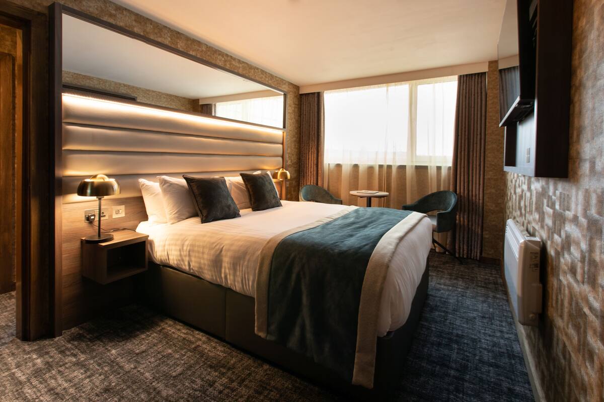 Quarto de hotel moderno com uma cama grande, mesas de cabeceira, uma pequena escrivaninha e uma cadeira.