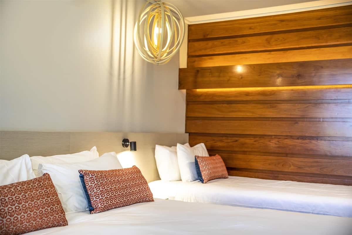 Quarto de hotel com cabeceira de madeira, cama grande com roupa de cama branca e almofadas estampadas, e luminária pendente moderna.