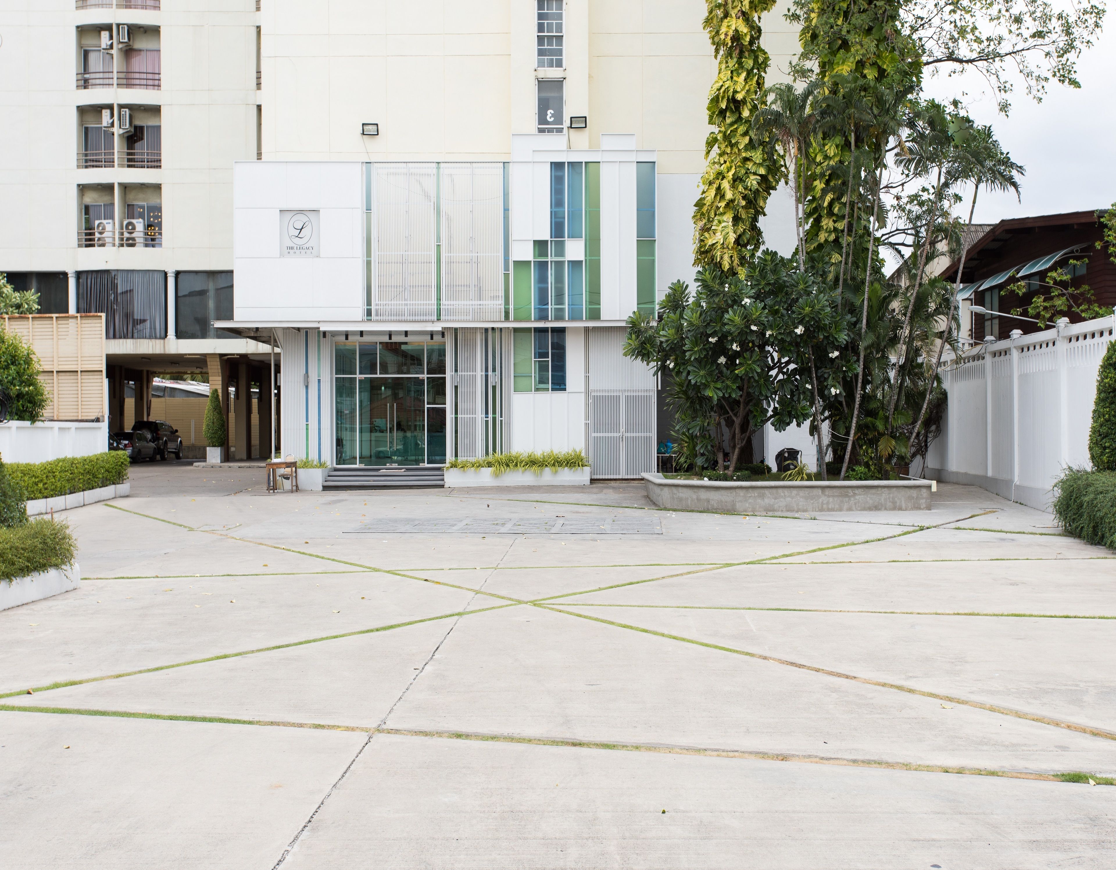Um edifício moderno com entrada de vidro, rodeado por um espaço ajardinado com pátio pavimentado.