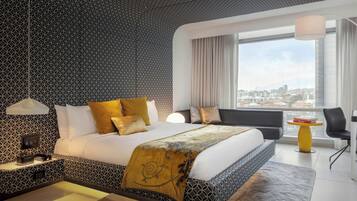 Quarto de hotel moderno com uma cama grande, vista para a cidade, uma mesinha e um sofá.
