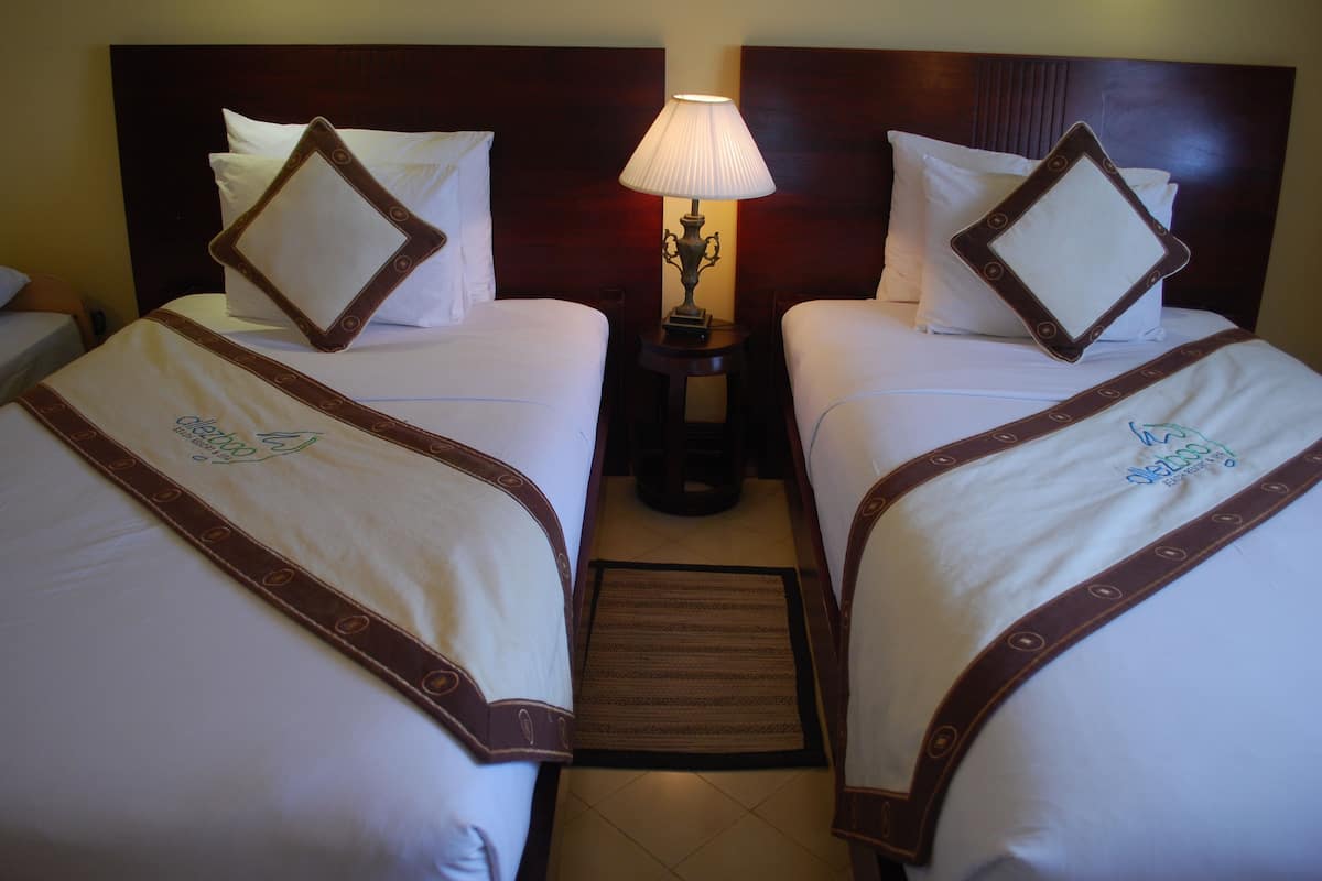 Quarto de hotel com duas camas, uma mesa de cabeceira com abajur e um tapete pequeno.
