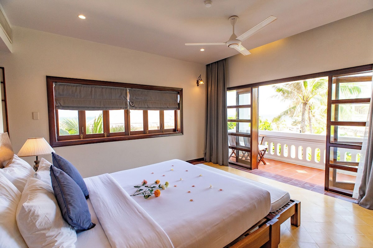 Quarto de hotel com cama, ventilador de teto, janela com vista para palmeiras e varanda com uma cadeira.