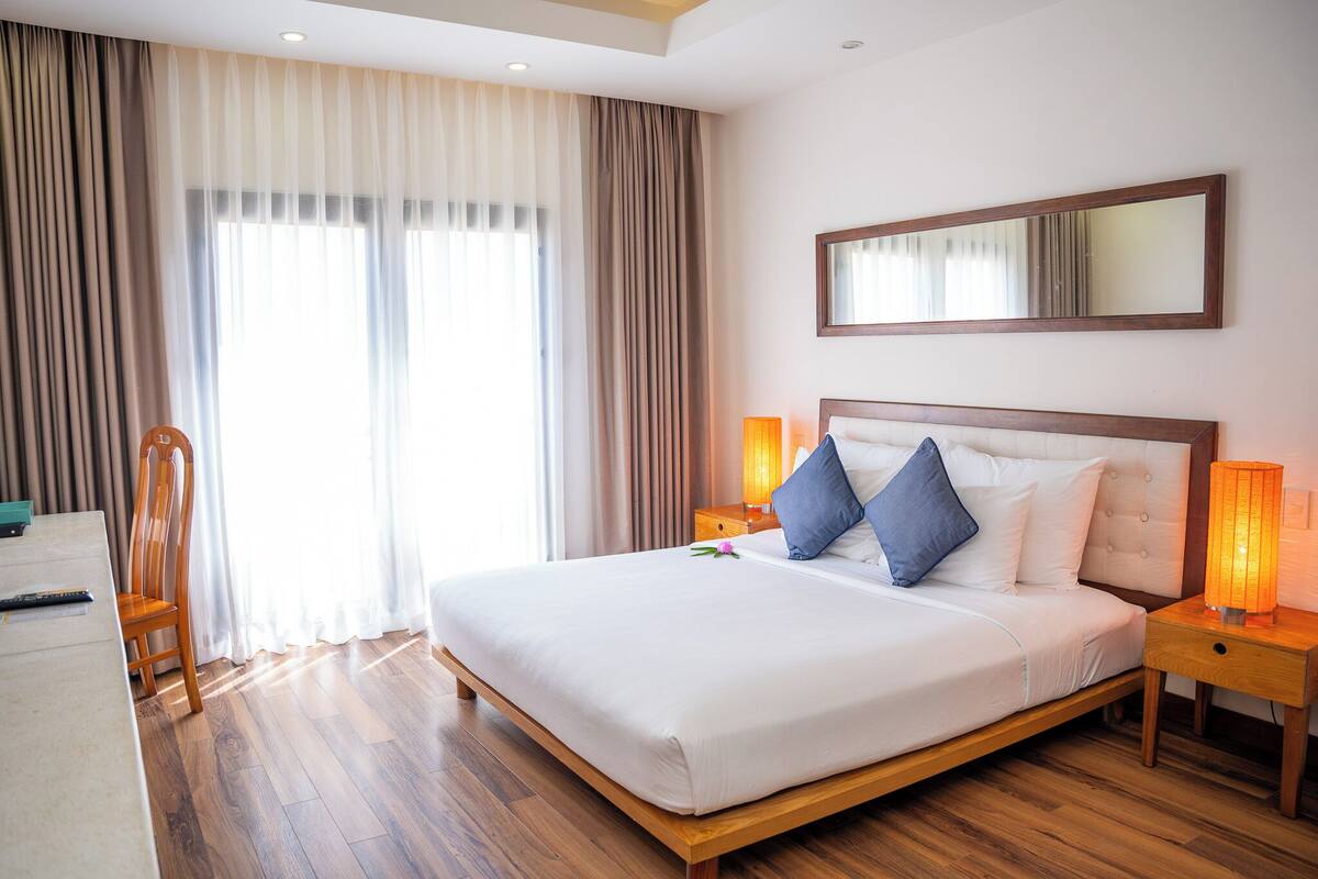 Quarto de hotel com uma cama grande, cabeceira de madeira, duas mesas de cabeceira com luminárias, uma cadeira de madeira e uma janela ampla com cortinas.