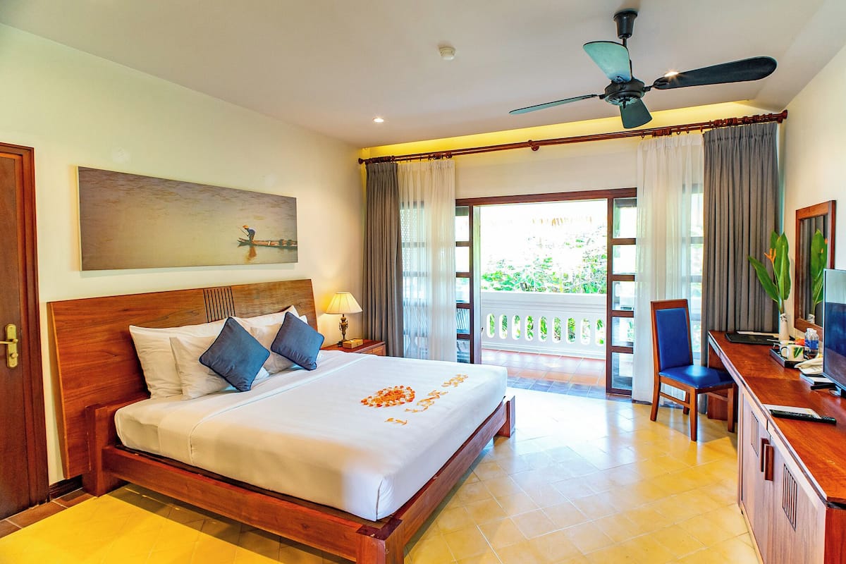 Quarto de hotel com uma cama grande, uma escrivaninha com televisão, um ventilador de teto e uma varanda com cortinas.