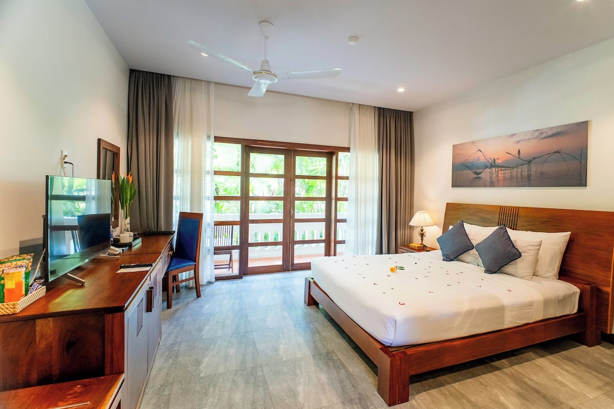 Quarto de hotel com cama, escrivaninha, cadeira, televisão e uma varanda com cortinas.