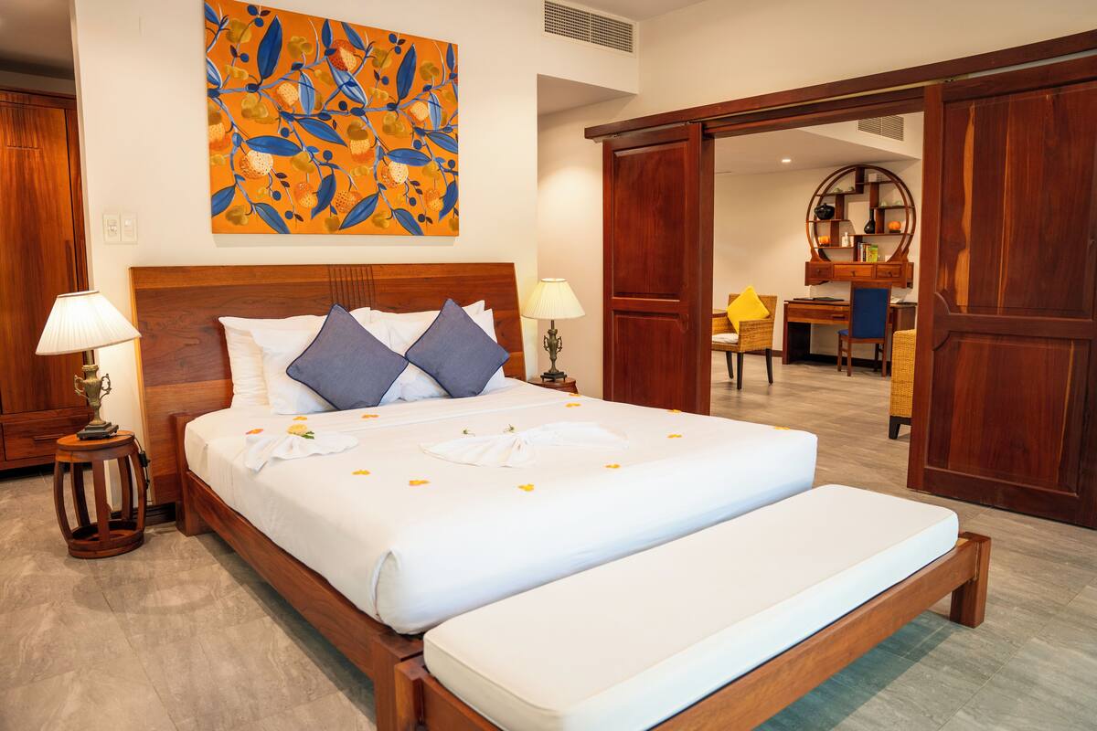 Uma cama bem arrumada com cabeceira de madeira, mesa de cabeceira com abajur e uma grande obra de arte floral na parede.
