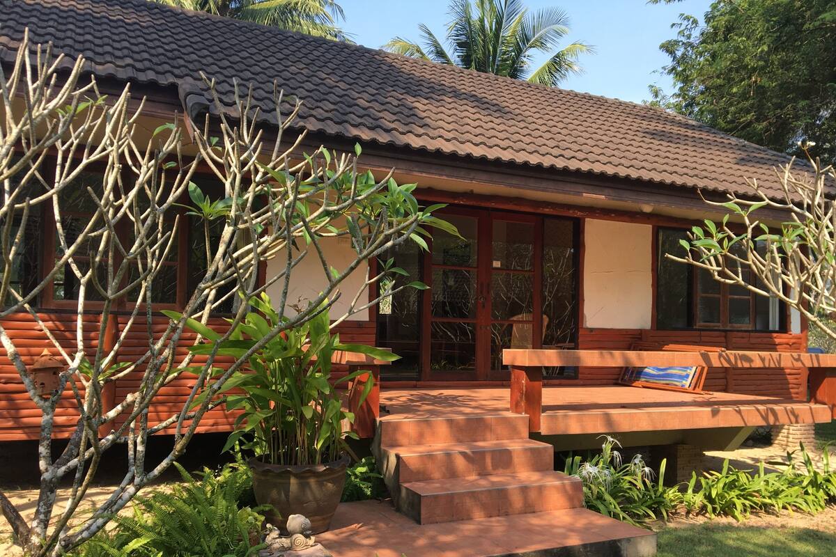 Uma casa de madeira com telhado de telhas, varanda com um banco e um vaso grande com planta.