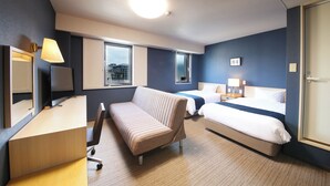 Quarto de hotel com escrivaninha, duas camas, um sofá, uma janela grande e vista para a cidade.